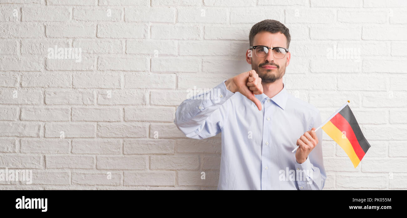 Young adult man over brick wall holding flag of Germany with angry face ...