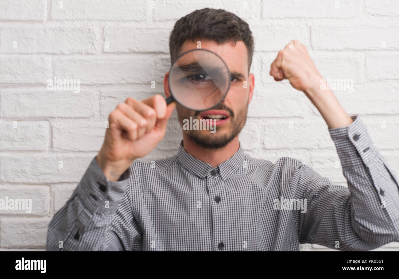 Young adult man over brick wall using magnifying glass annoyed and ...