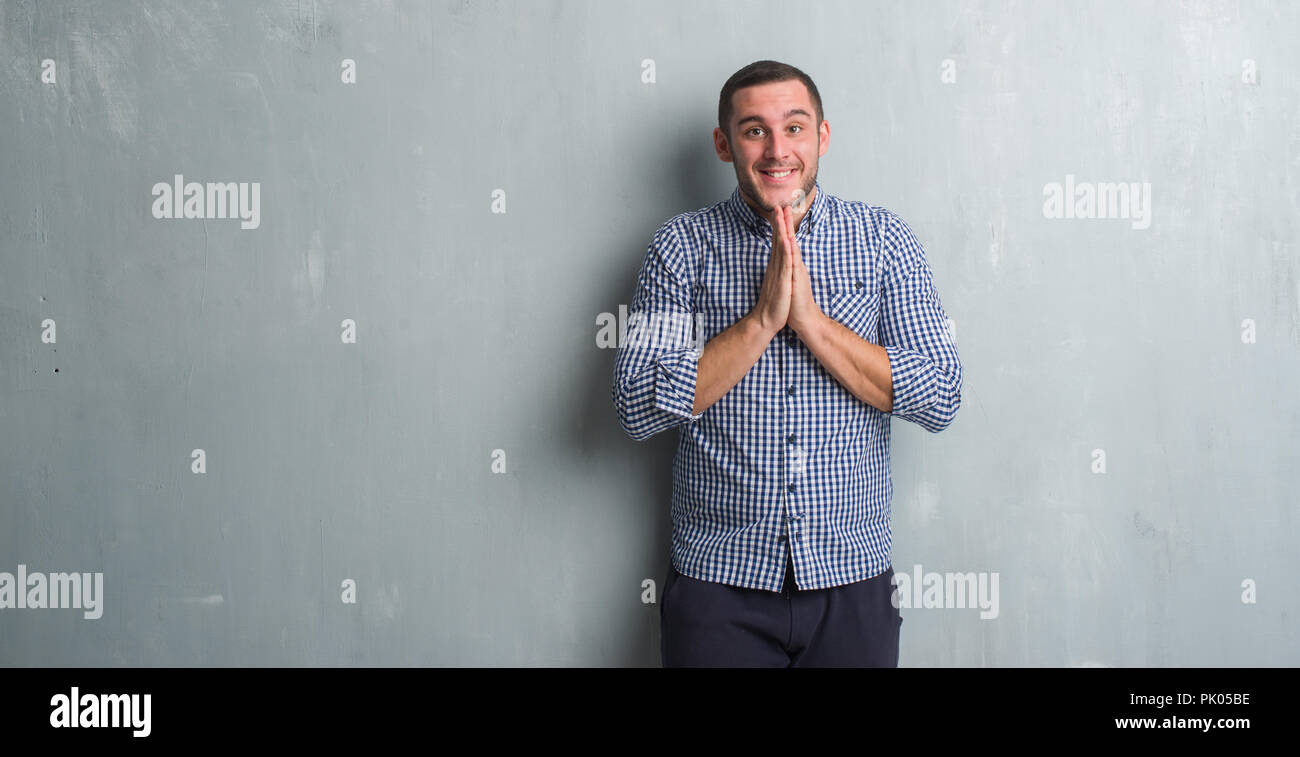 Young caucasian man over grey grunge wall praying with hands together ...