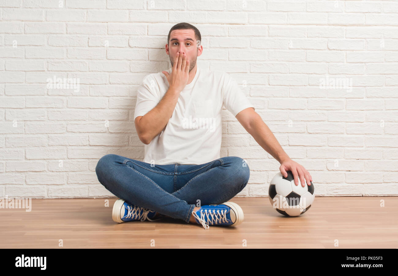 Young caucasian man sitting over white brick wall holding soccer ...