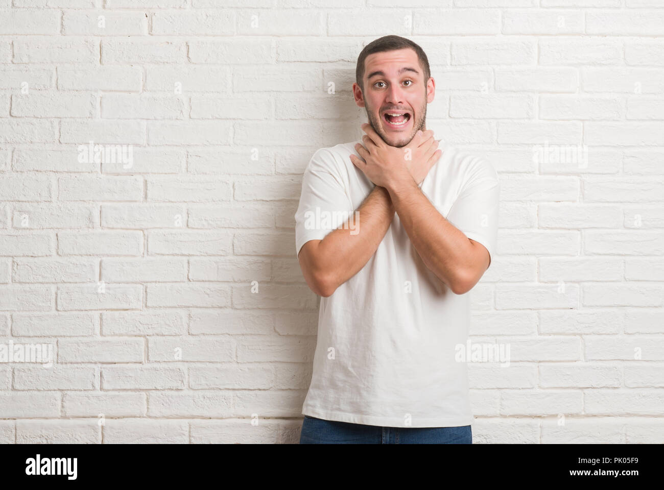 Young caucasian man standing over white brick wall smelling something ...