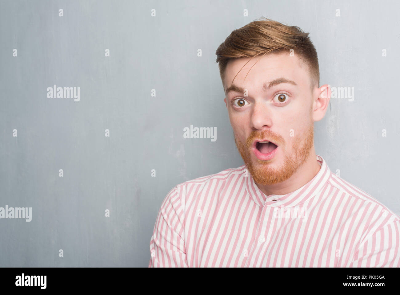 Young redhead man over grey grunge wall scared in shock with a surprise ...
