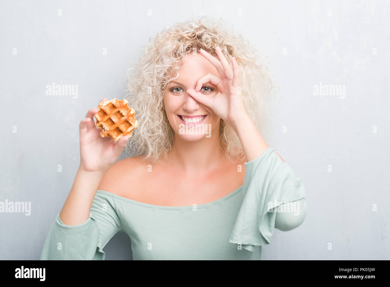 Young blonde woman over grunge grey background eating belgian waffle ...