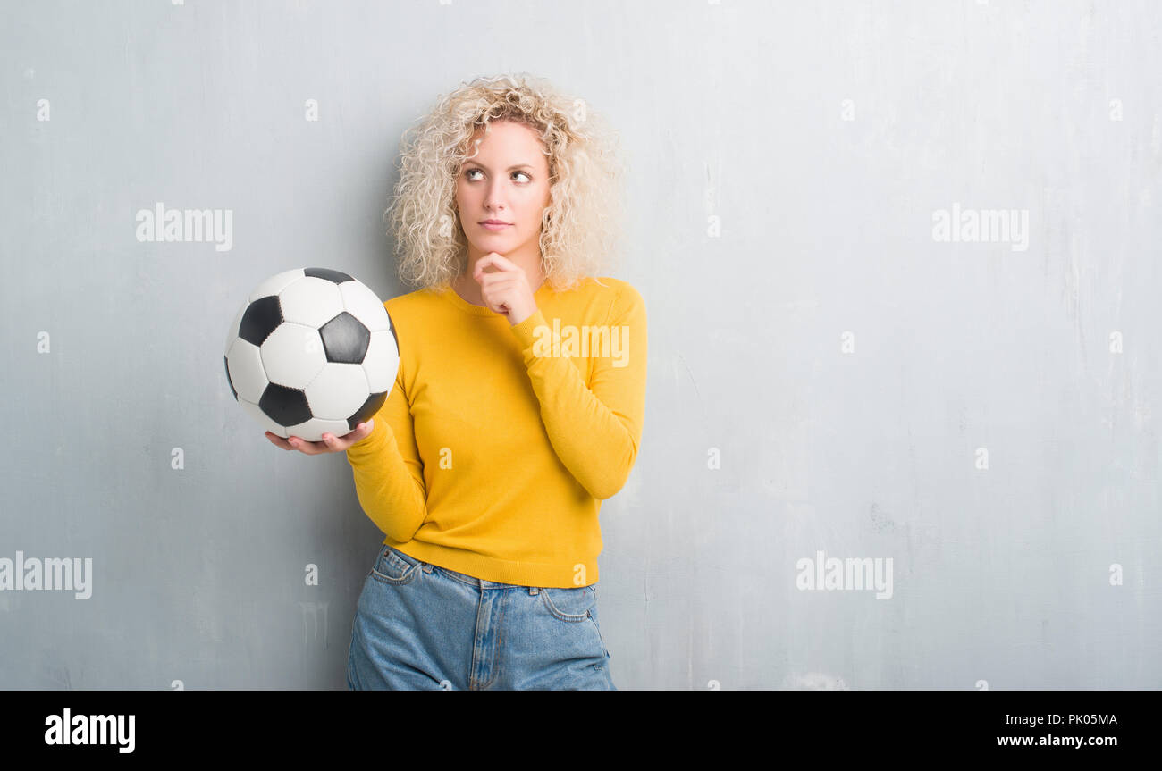 Young blonde woman over grunge grey background holding soccer football ...