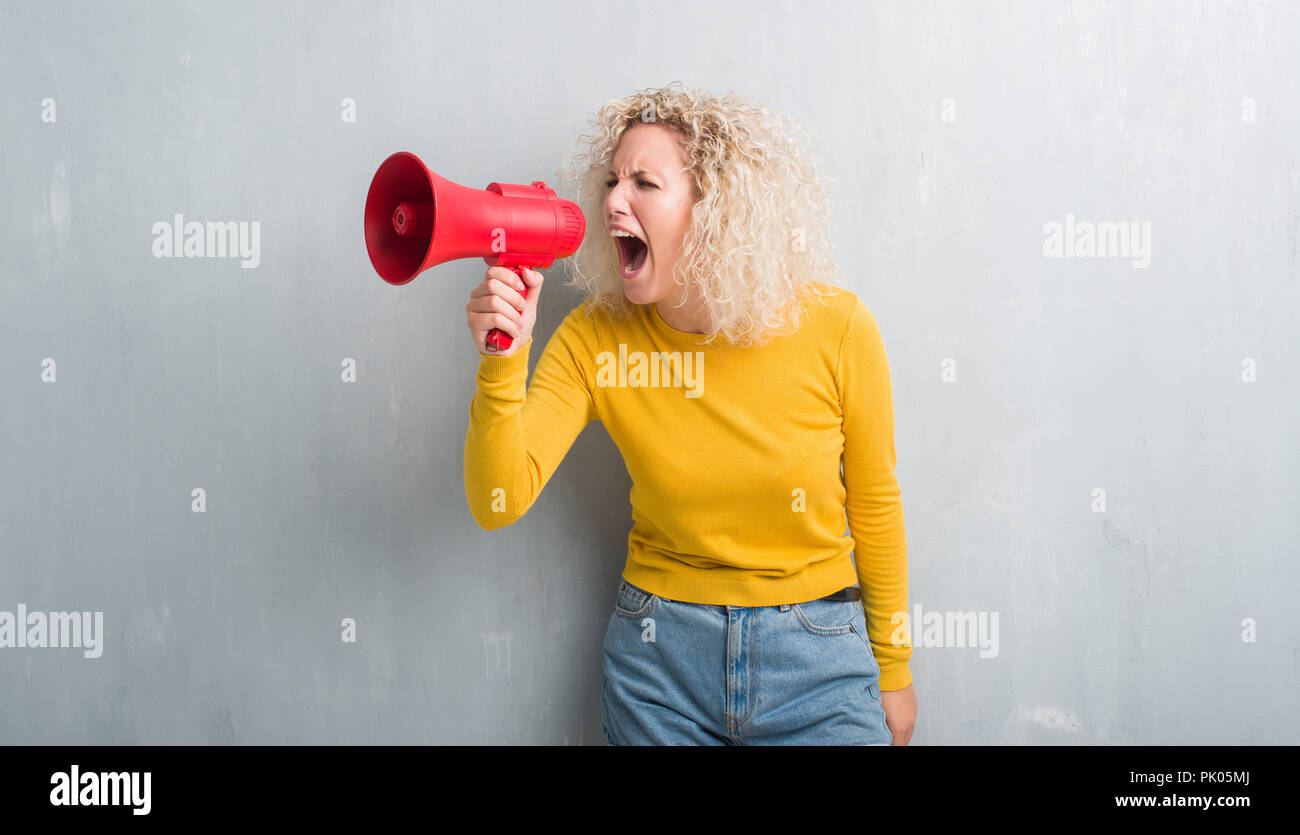 Young blonde woman over grunge grey background holding megaphone ...