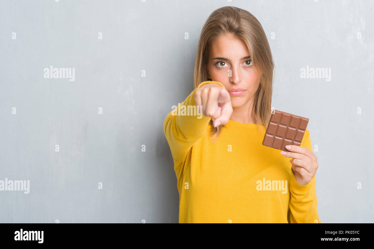 Beautiful young woman over grunge grey wall eating chocolate bar ...