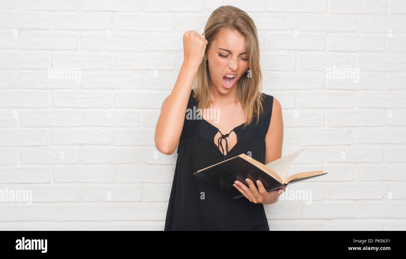 Beautiful young woman over white brick wall reading a book annoyed and ...