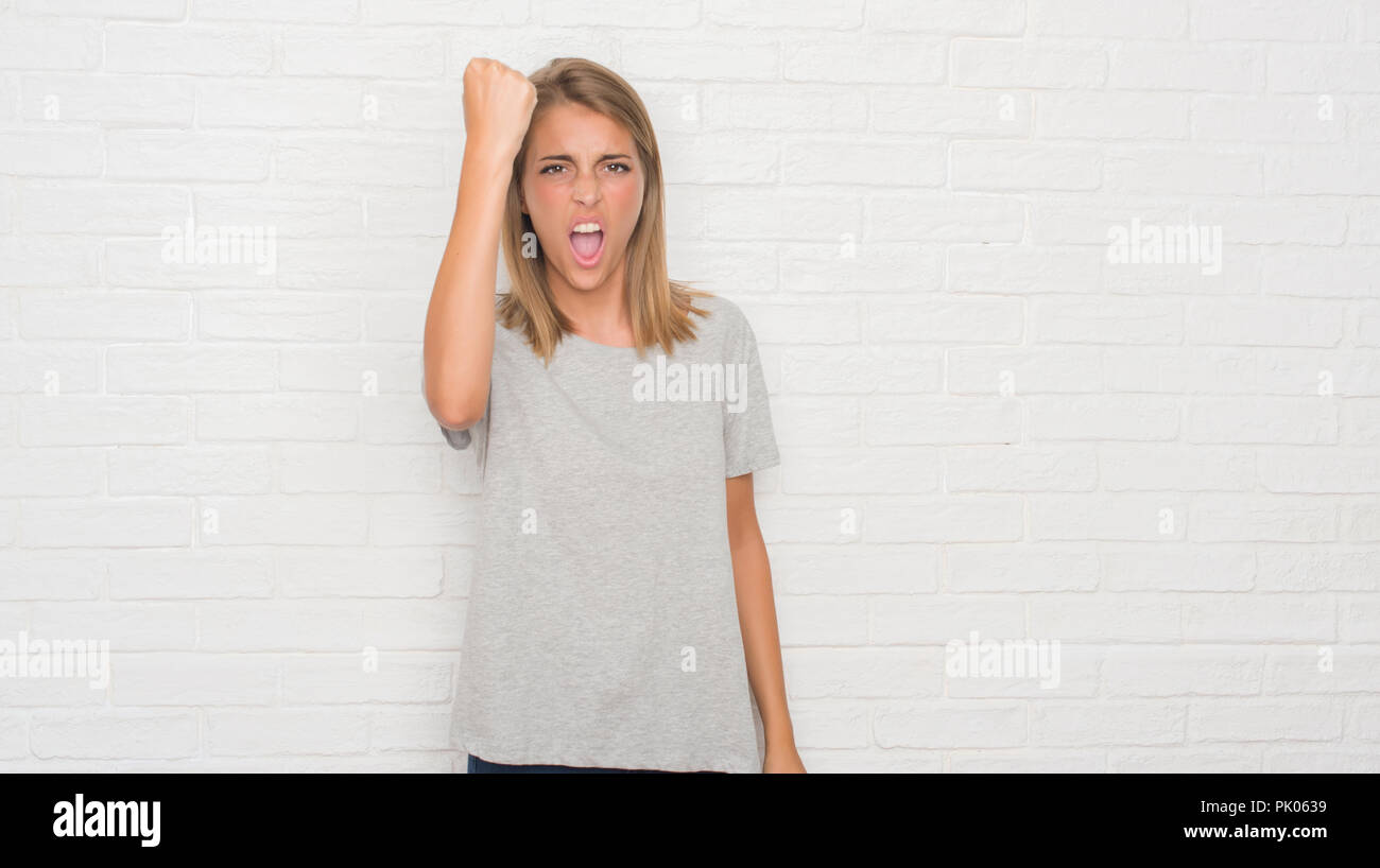Beautiful young woman over white brick wall angry and mad raising fist ...