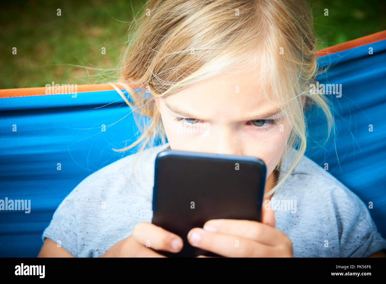 Child blond girl playing with a smart phone while lying on hammock ...
