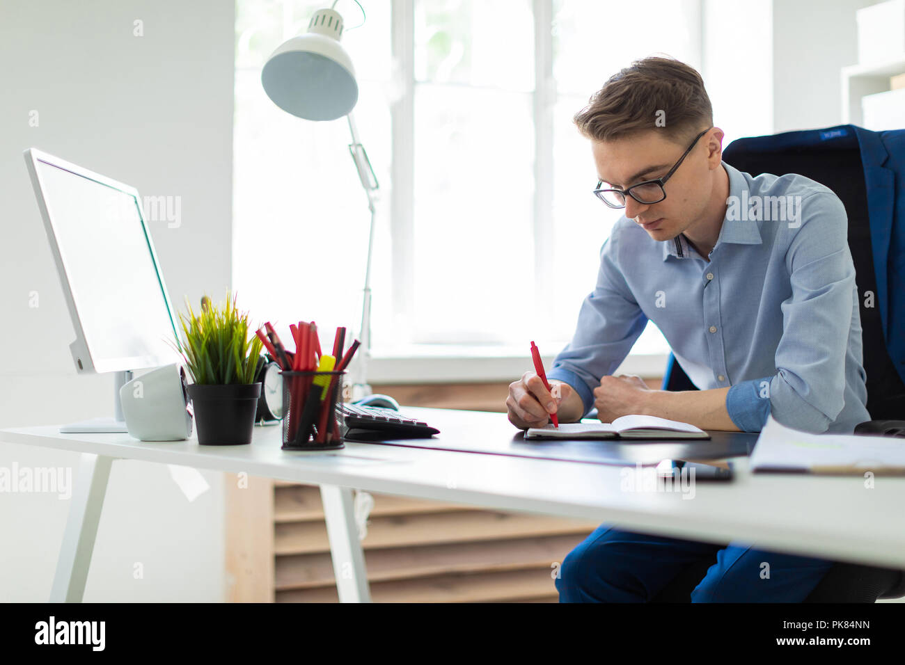 A young man sits in the office at a computer desk and writes in a ...