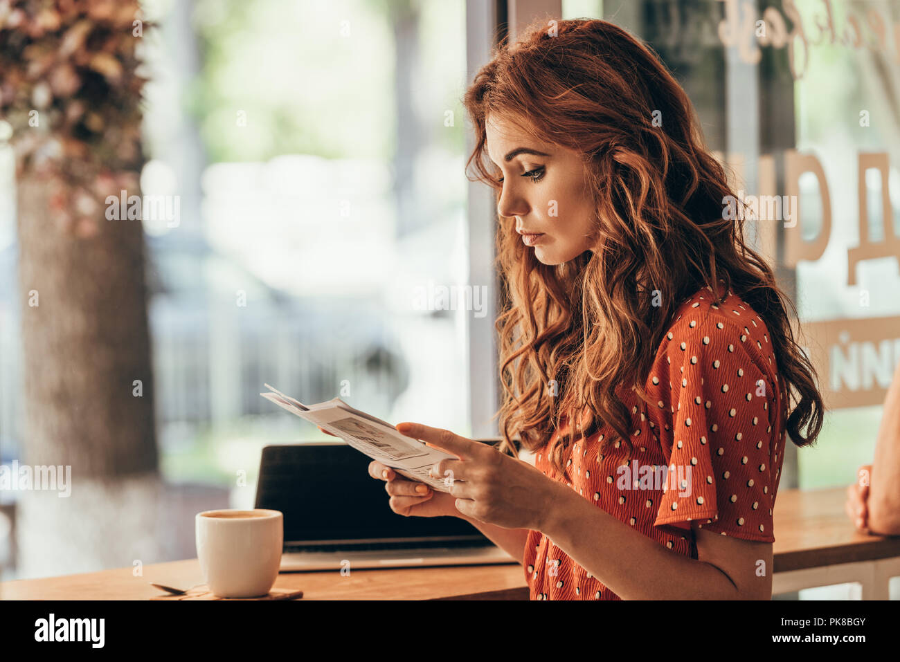 side view of young woman reading newspaper at table with laptop in ...