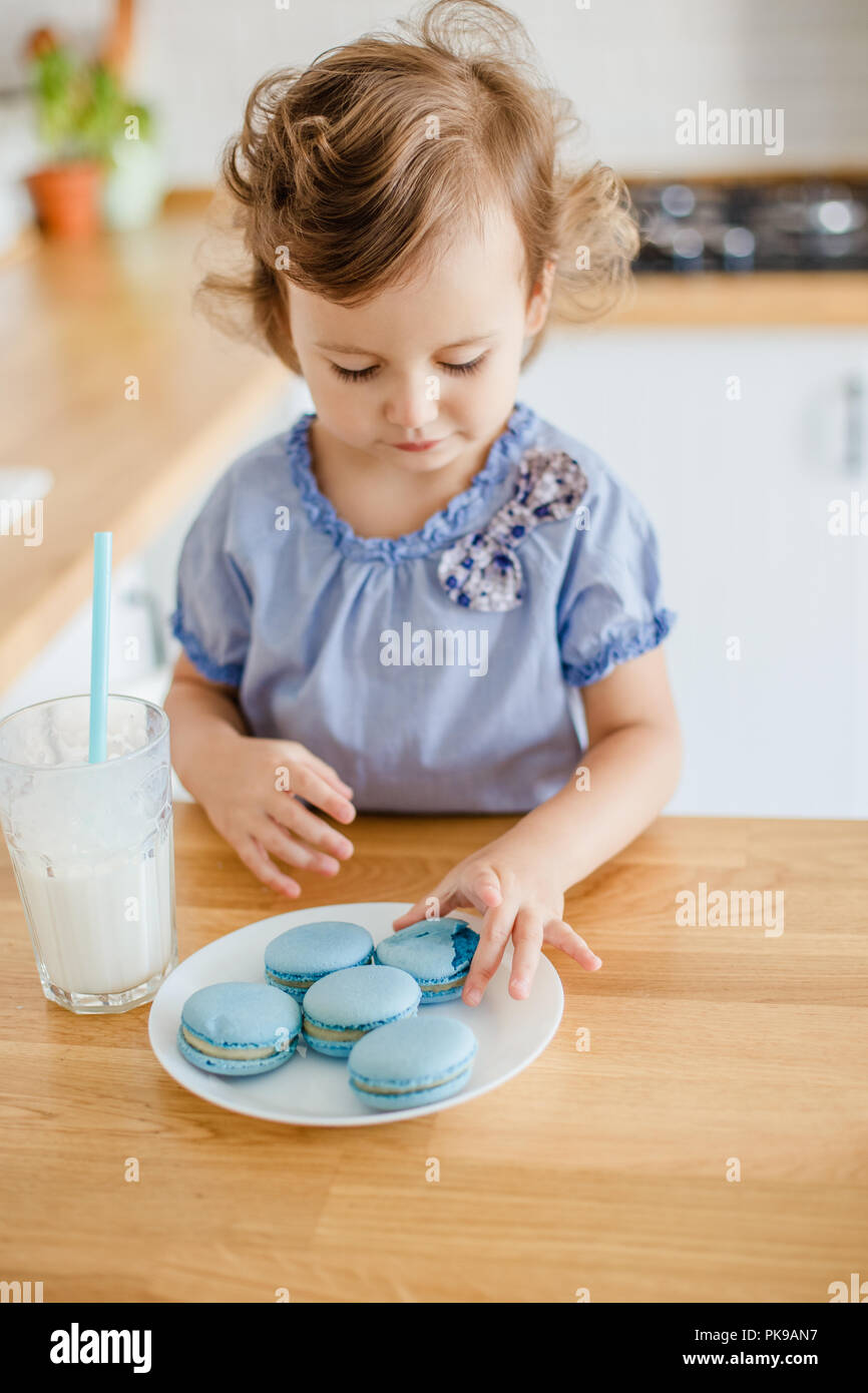 Adorable little girl eating blue macaroons on the white modern kitchen ...