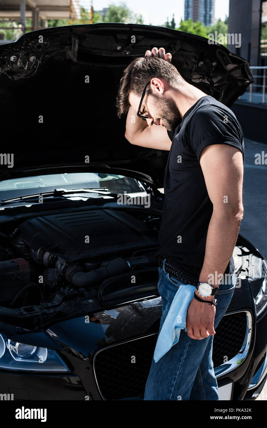 side view of confused man scratching head near broken car engine at ...