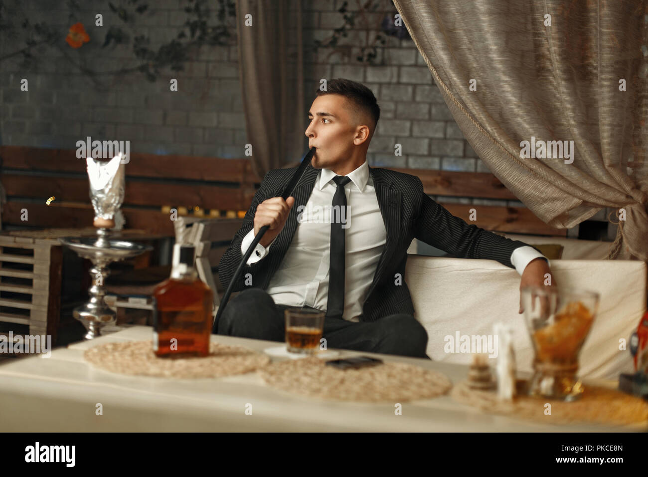 Young stylish man in a suit rests in the bar. Man drinking whiskey and smoking a hookah Stock ...