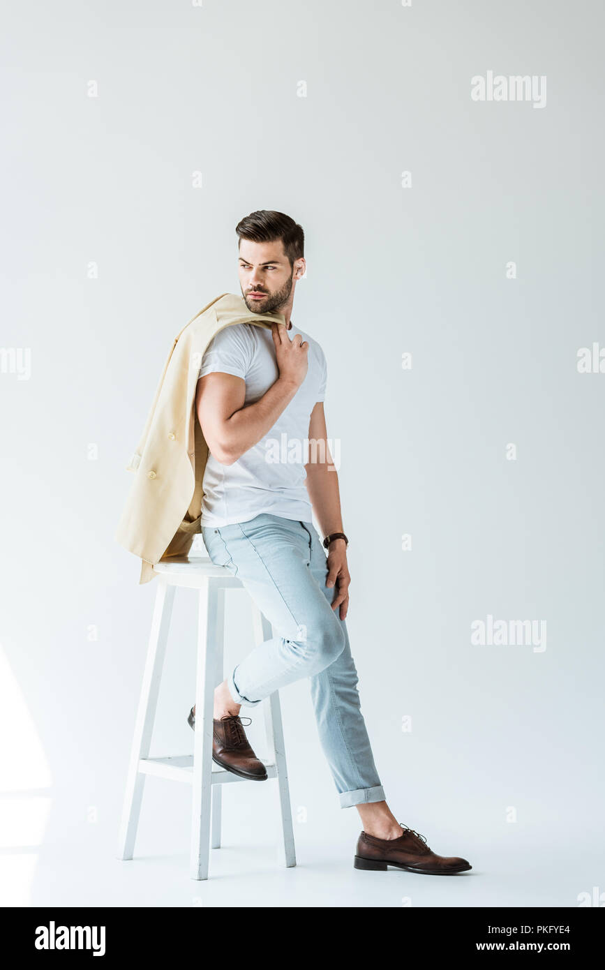 Stylish young man holding jacket and sitting on stool on white ...