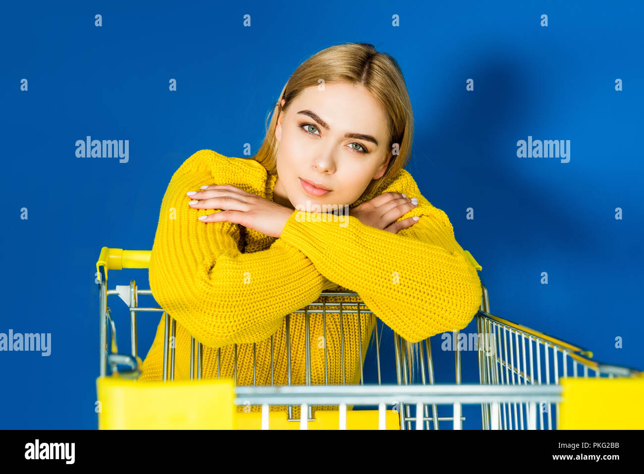 Elegant blonde woman in yellow garment leaning on shopping cart on blue ...