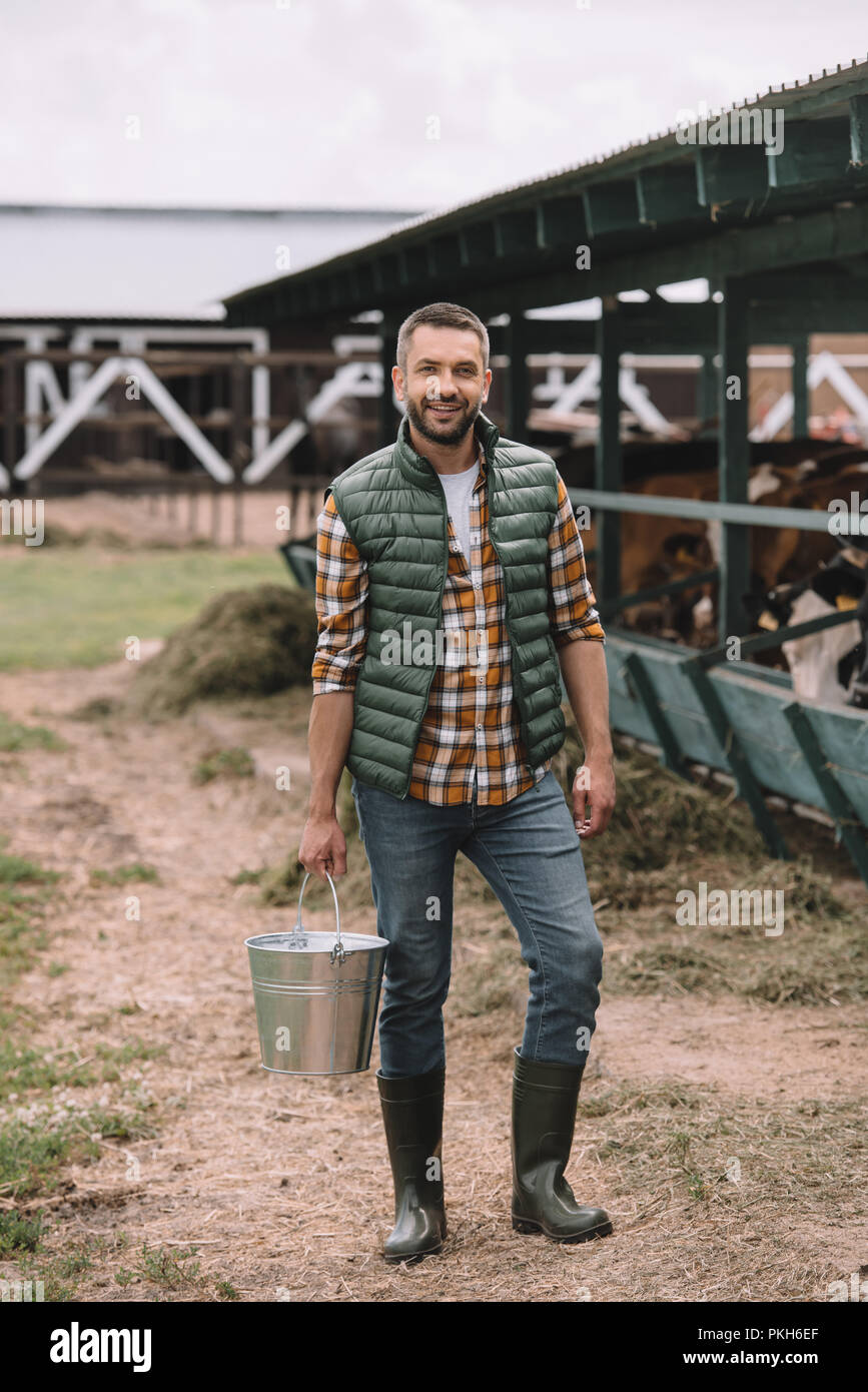 handsome male farmer holding bucket and smiling at camera at cowshed ...