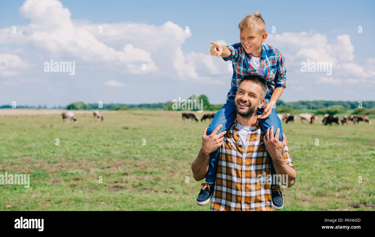 happy father carrying smiling son on neck and looking away in field ...