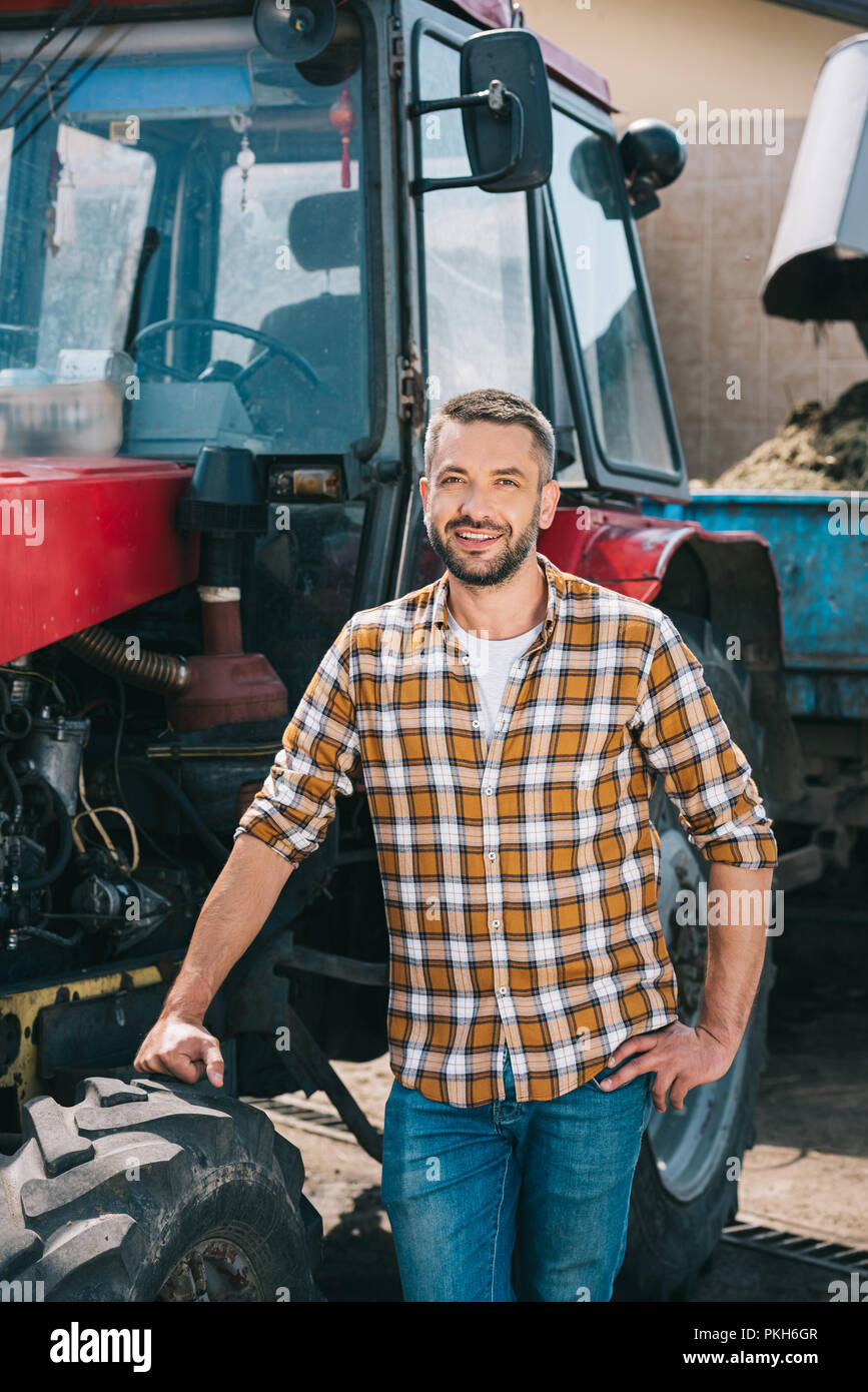 handsome middle aged farmer standing near tractor and smiling at camera ...