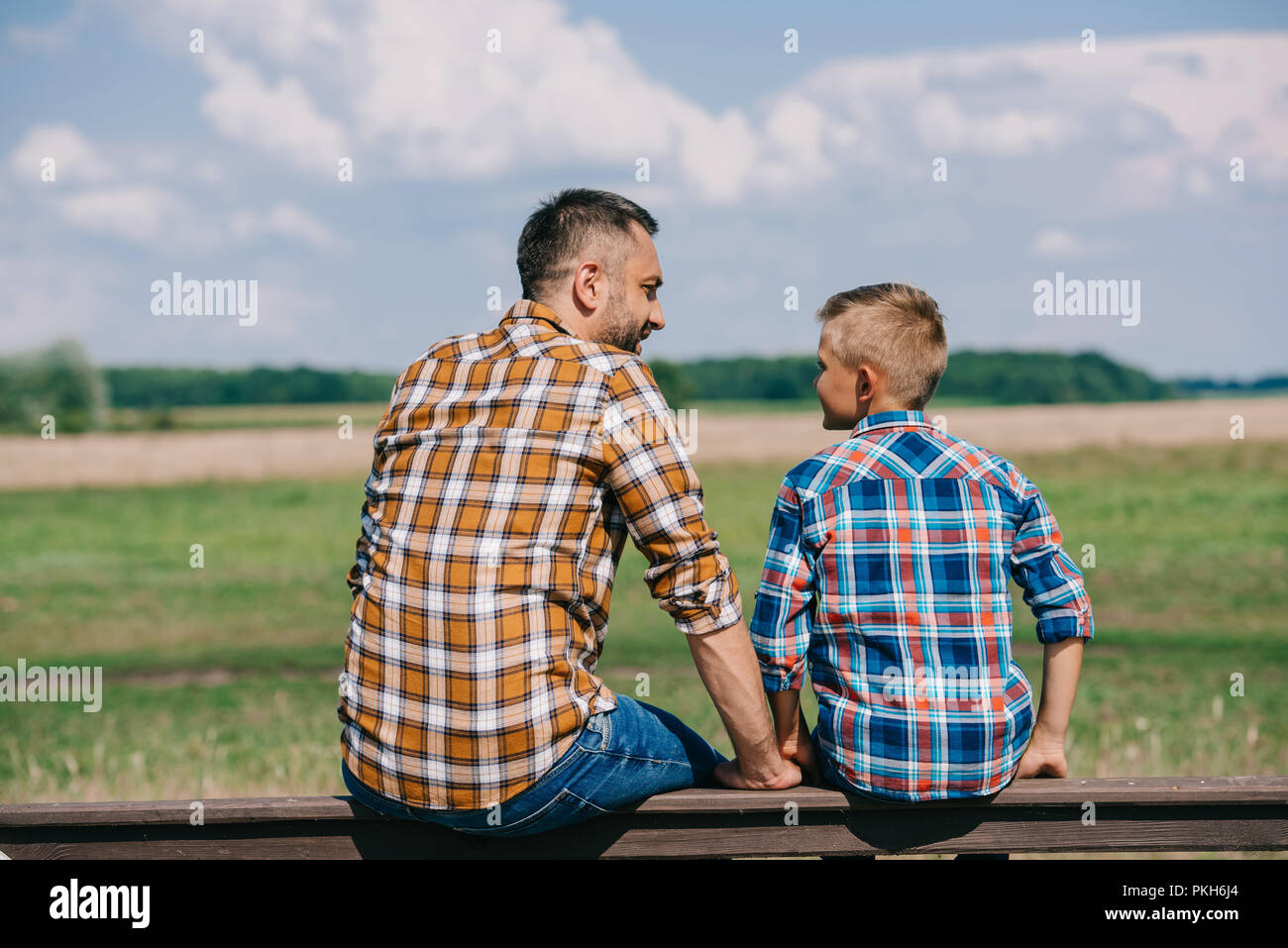 back view of father and son sitting on fence and smiling each other at ...