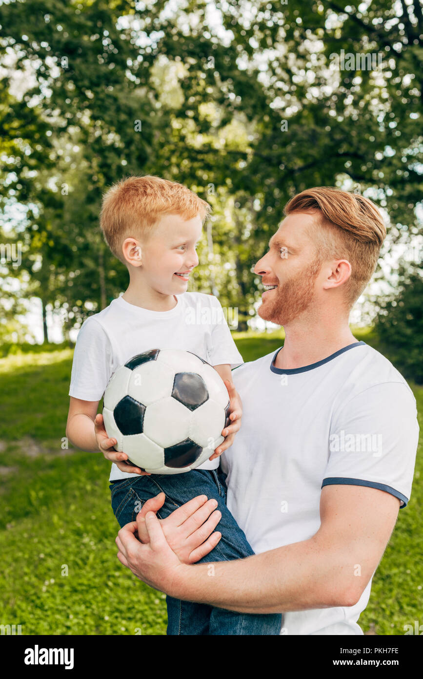 happy father and son holding soccer ball and smiling each other at park ...