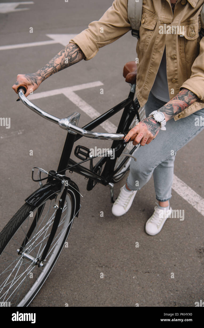 partial view of tattooed woman standing with bicycle at parking lot ...