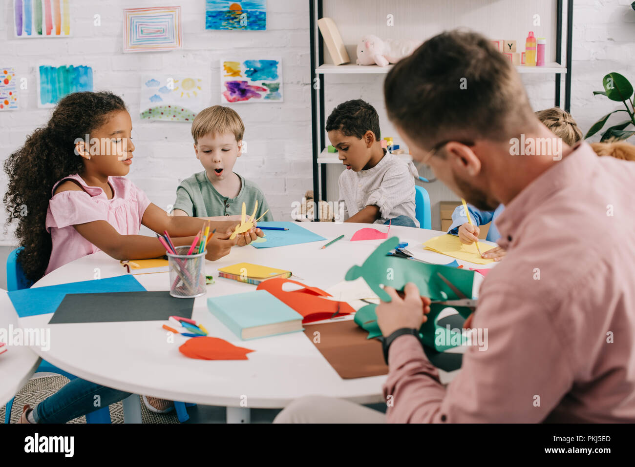 teacher and multiracial preschoolers with colorful papers and scissors ...