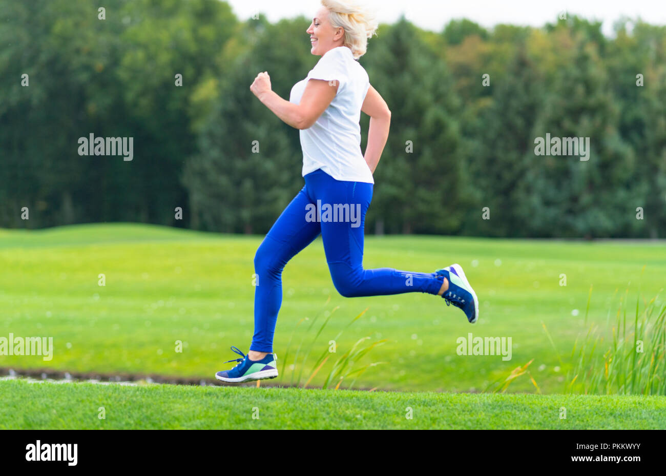Happy healthy mature woman running through a park at speed smiling to ...