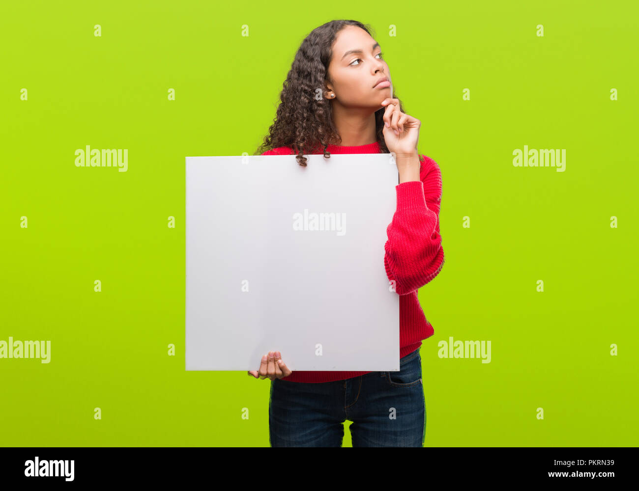 Young hispanic woman holding blank banner serious face thinking about ...