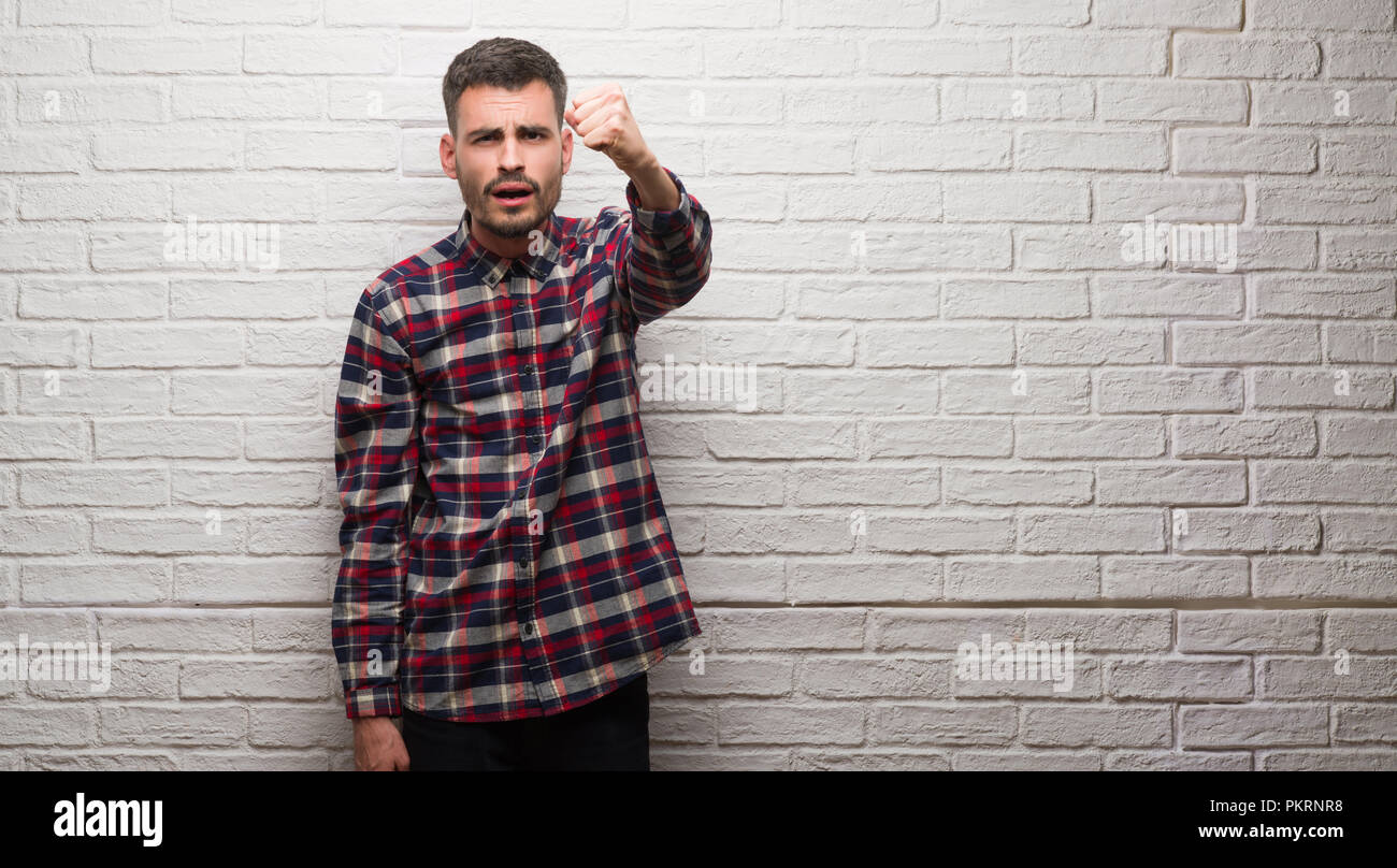 Young adult man over white brick wall annoyed and frustrated shouting ...