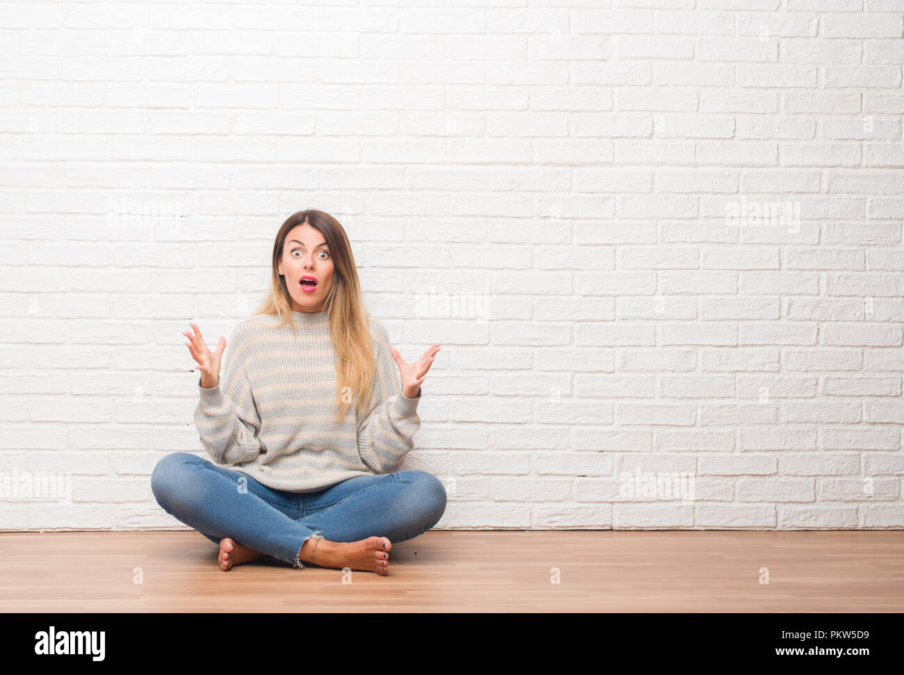 Young adult woman sitting on the floor over white brick wall at home ...
