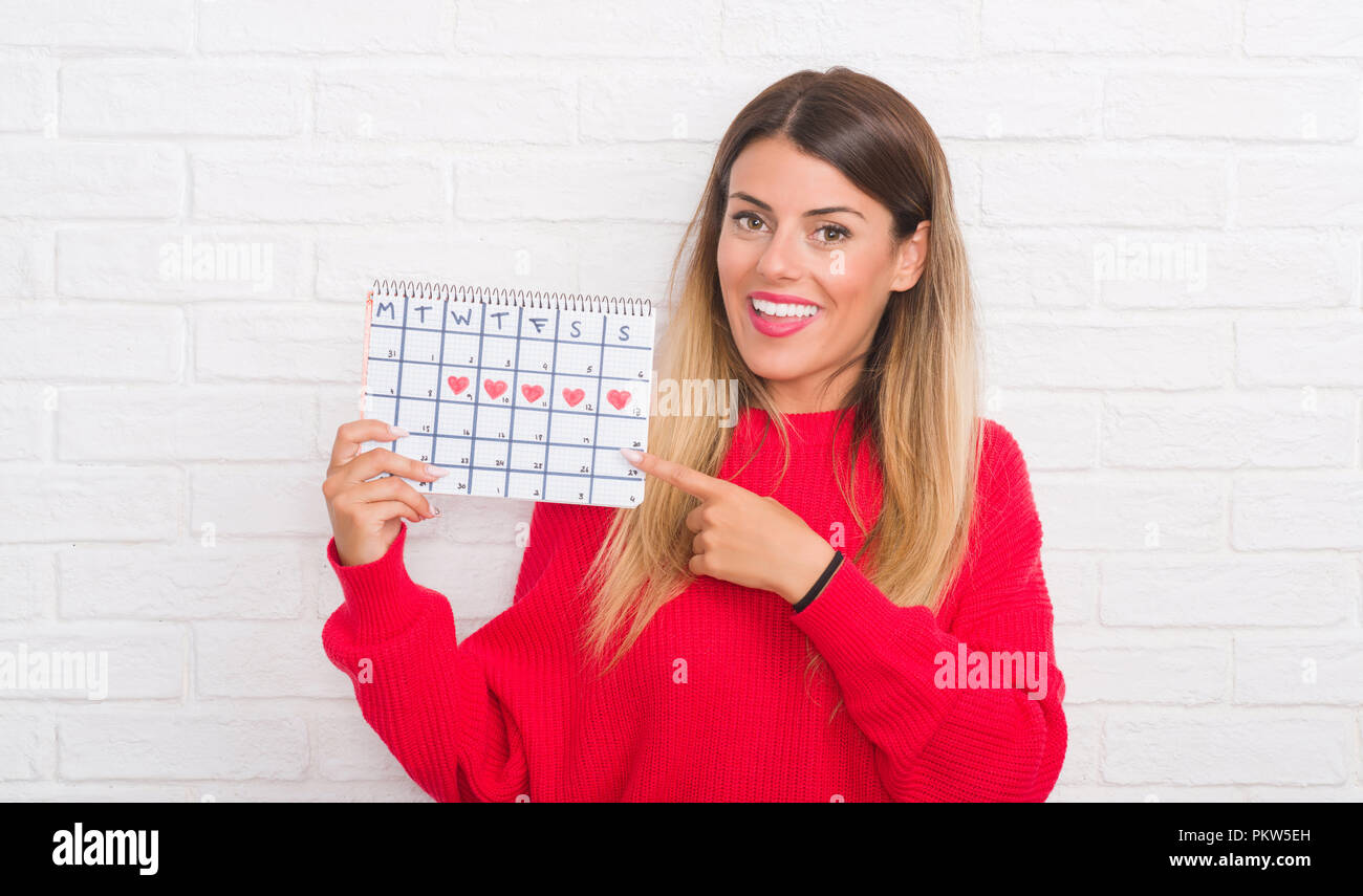 Young adult woman over white brick wall holding period calendar very ...