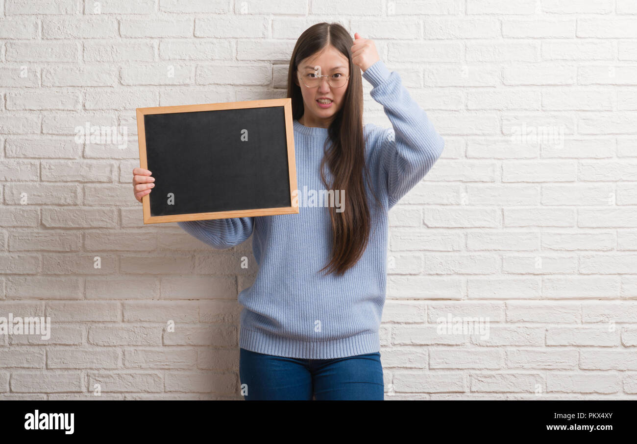 Young Chinese woman over brick wall holding blackboard annoyed and ...