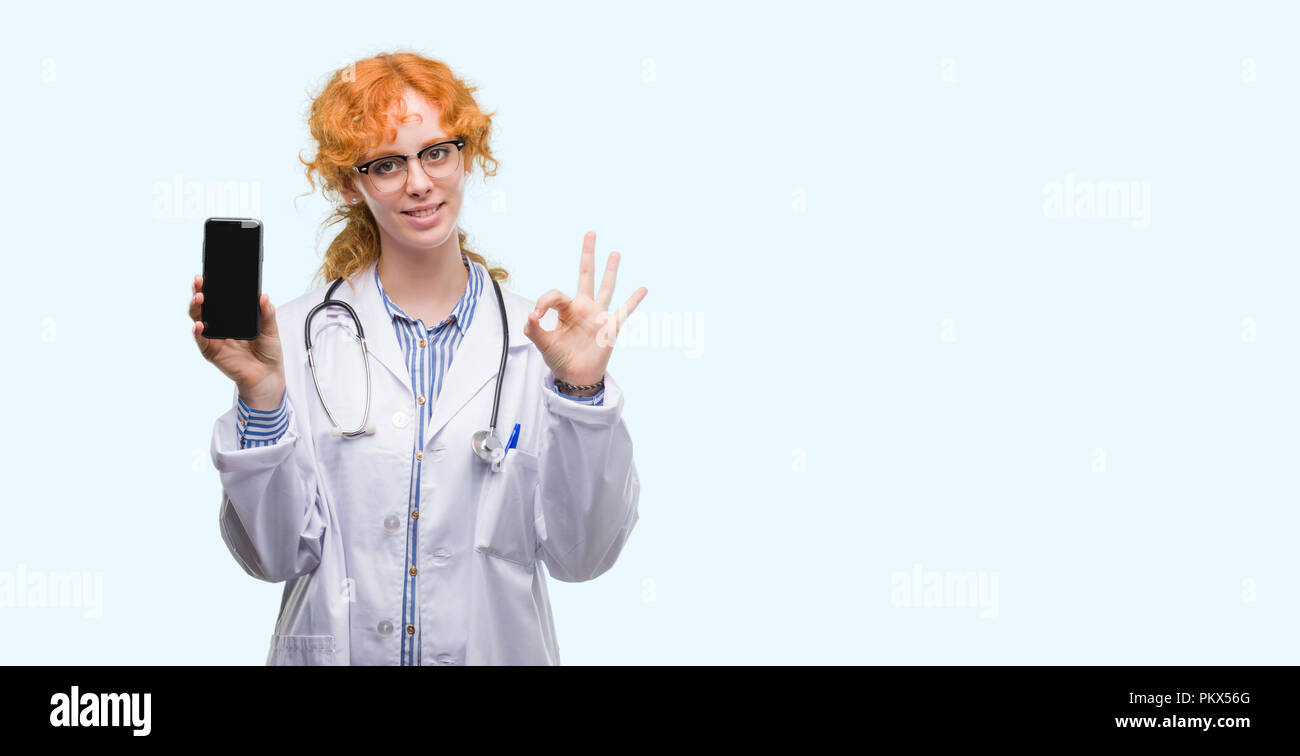 Young redhead doctor woman showing smartphone doing ok sign with ...