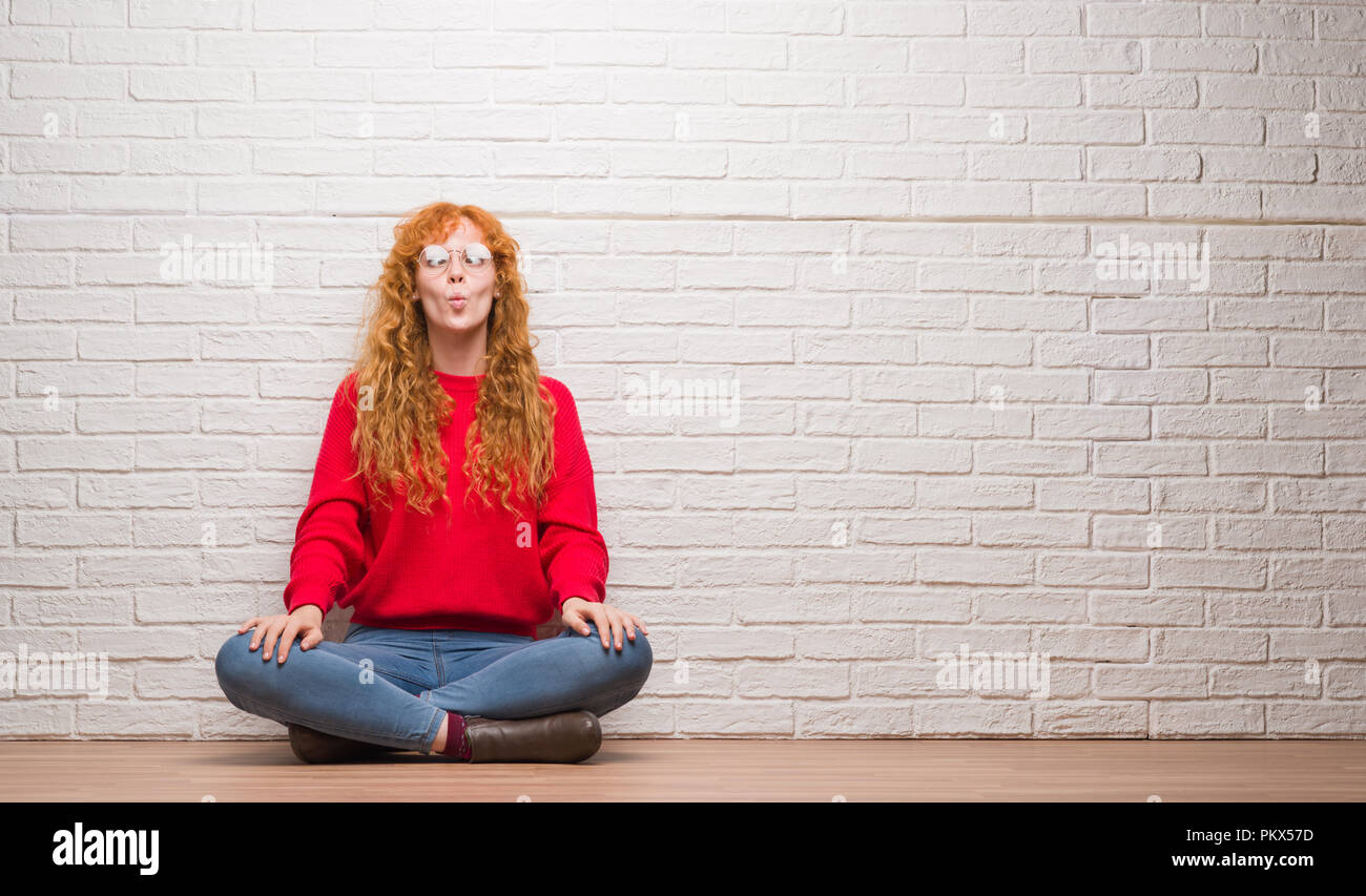Young redhead woman sitting over brick wall making fish face with lips ...