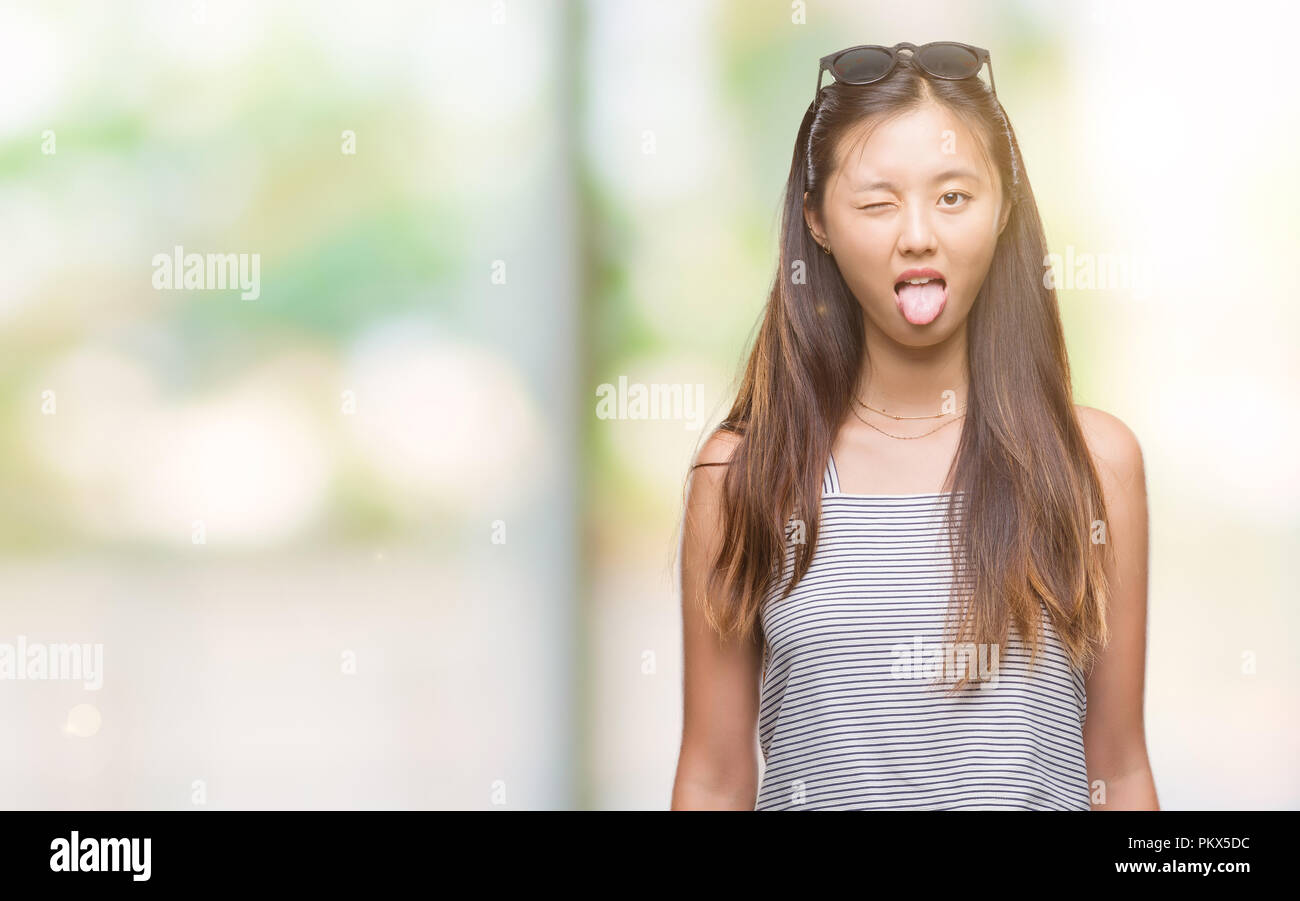 Young asian woman wearing sunglasses over isolated background sticking tongue out happy with ...