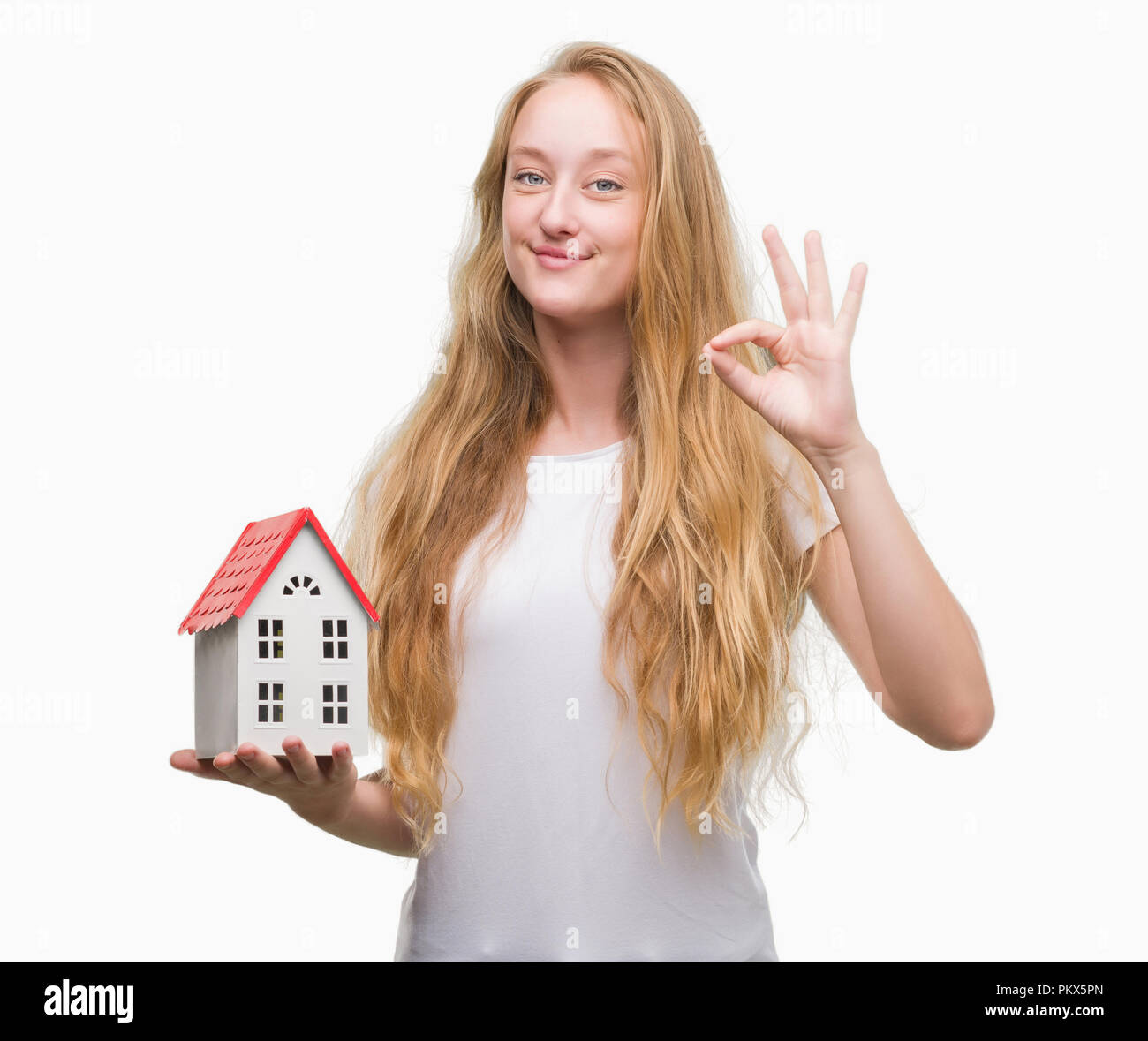 Blonde teenager woman holding family house doing ok sign with fingers ...