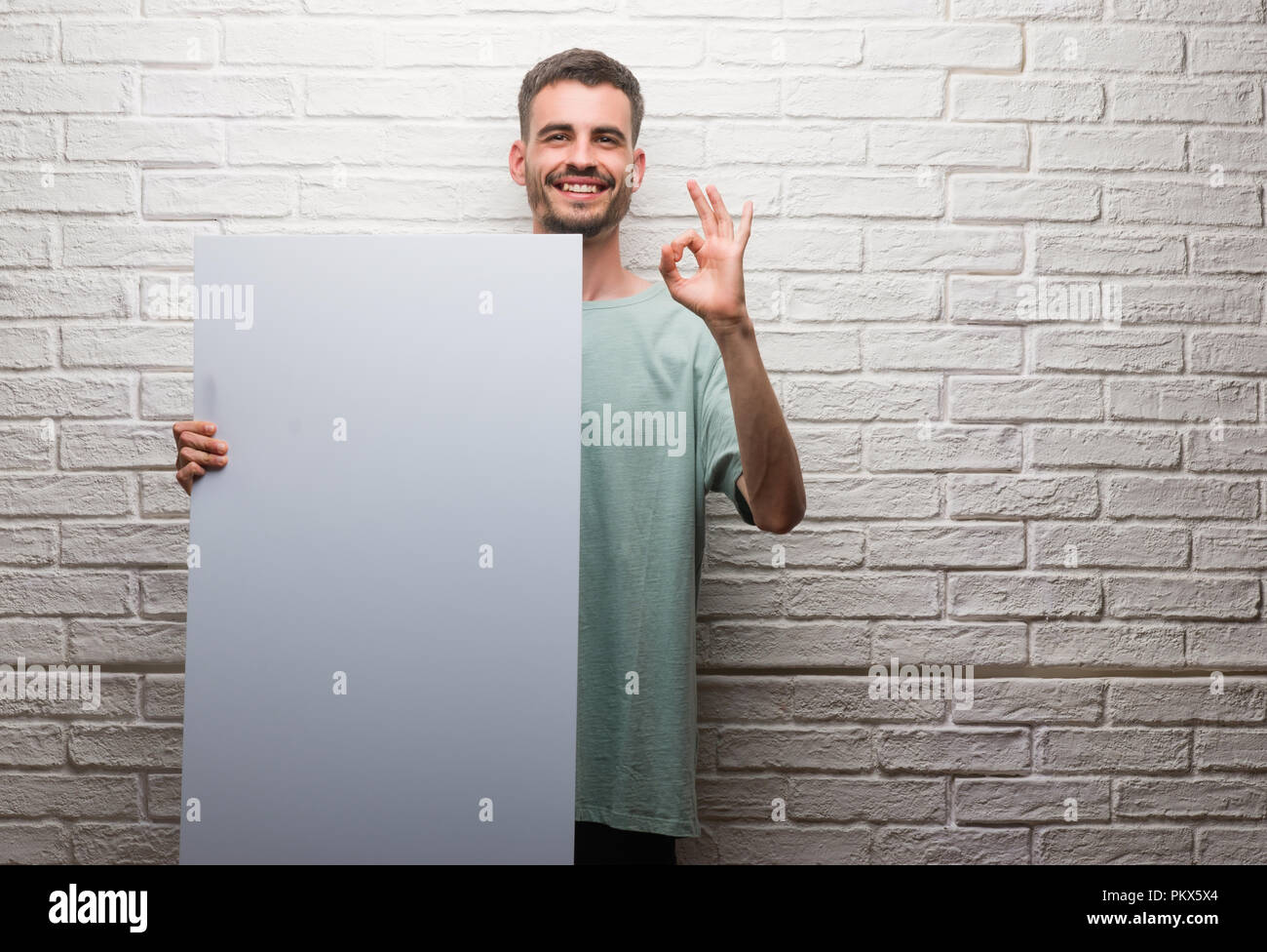 Young adult man over brick wall holding banner doing ok sign with ...