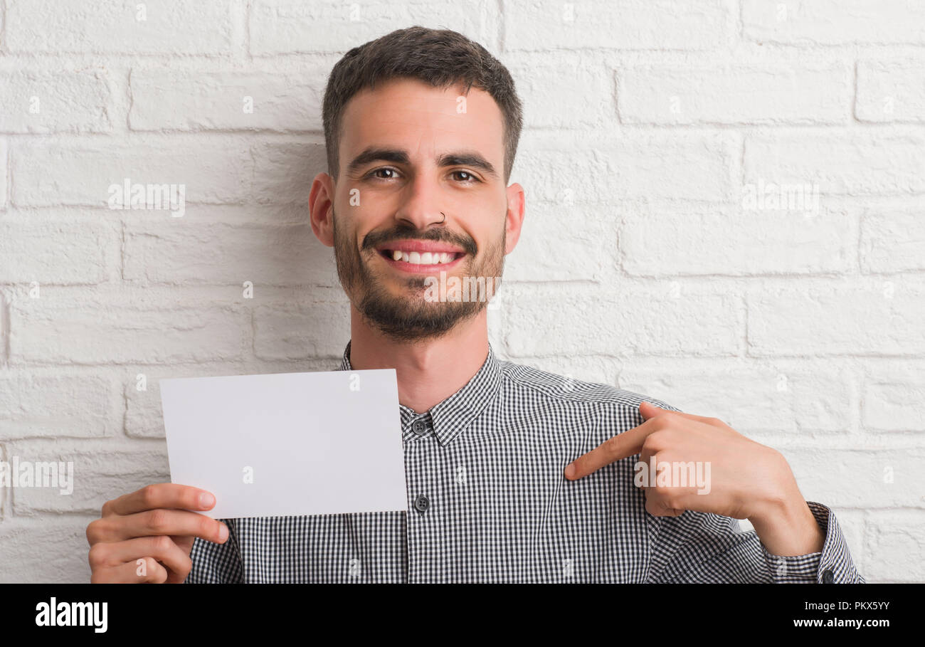 Young adult man over brick wall holding blank paper with surprise face ...