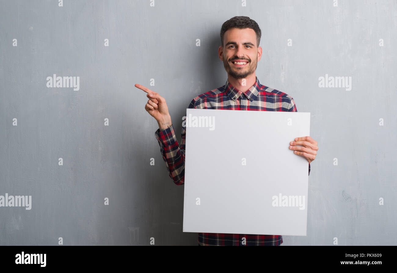 Young adult man over brick wall holding banner very happy pointing with ...