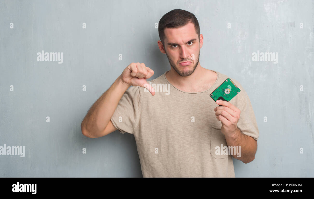 Young caucasian man over grey grunge wall holding hard drive with angry ...