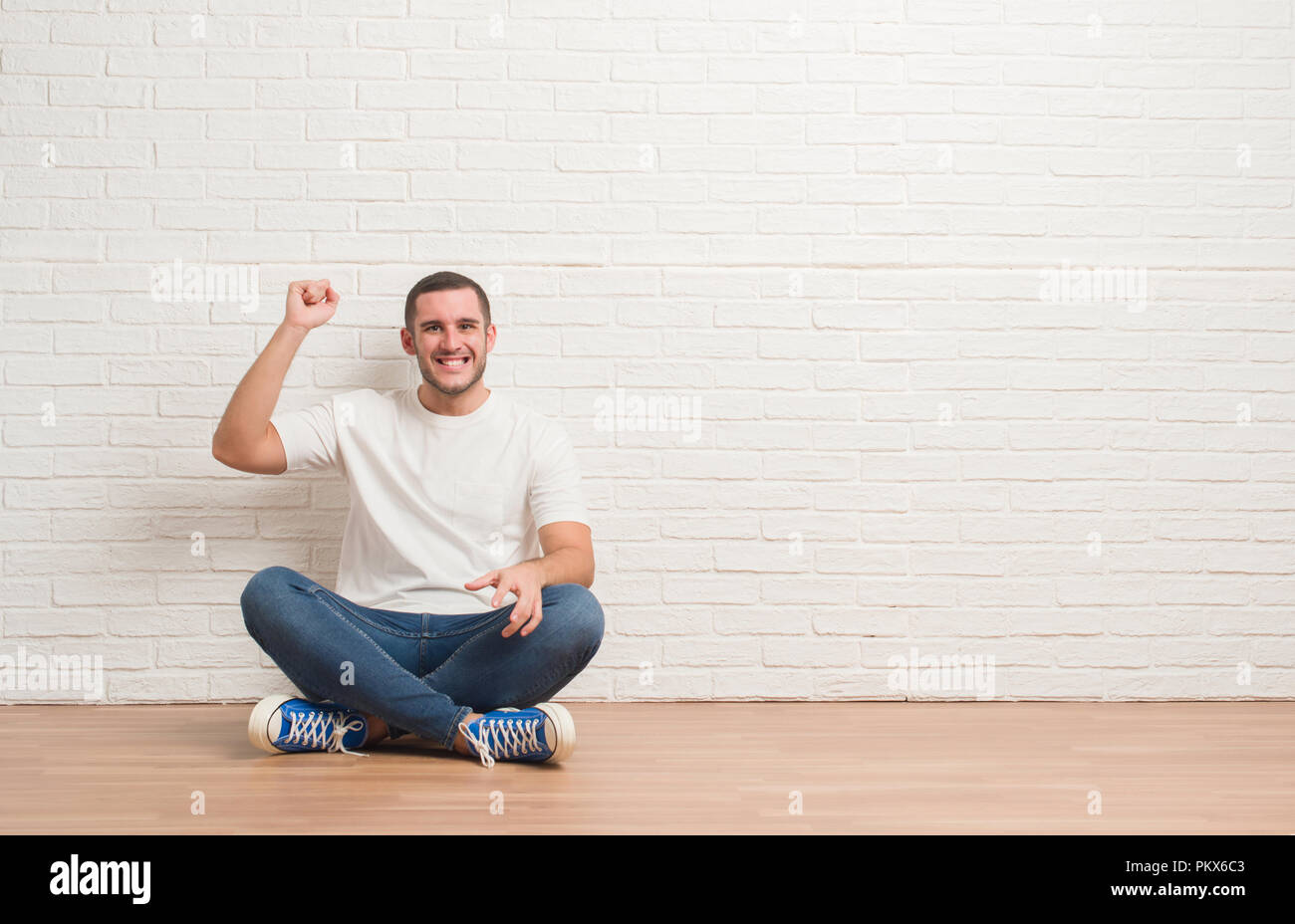 Young caucasian man sitting on the floor over white brick wall angry ...