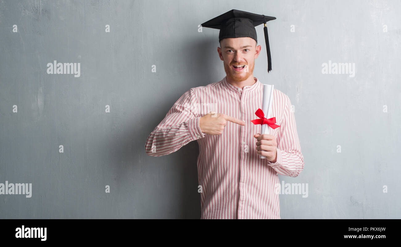Young redhead man over grey grunge wall wearing graduate cap holding ...