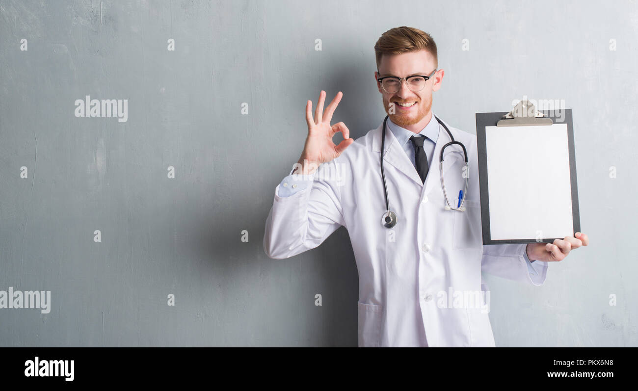 Young redhead doctor man over grey grunge wall holding clipboard doing ...