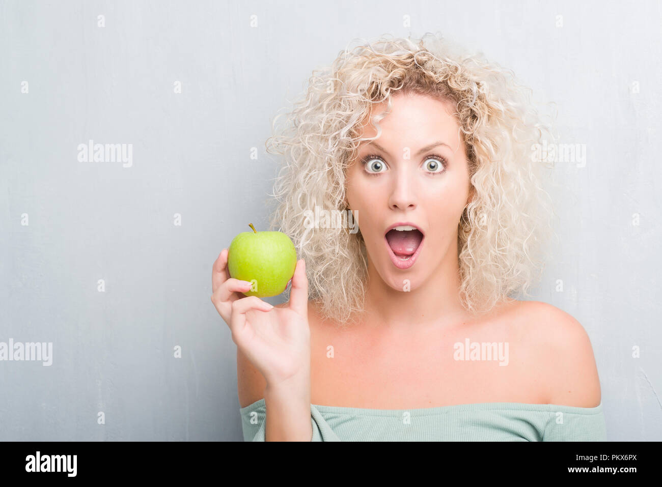 Young blonde woman over grunge grey background eating green apple ...