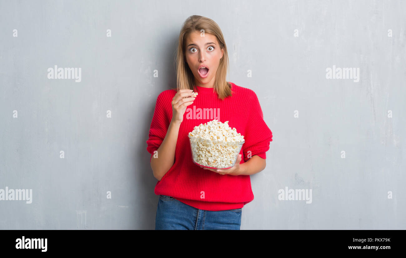 Beautiful young woman over grunge grey wall eating pop corn scared in ...