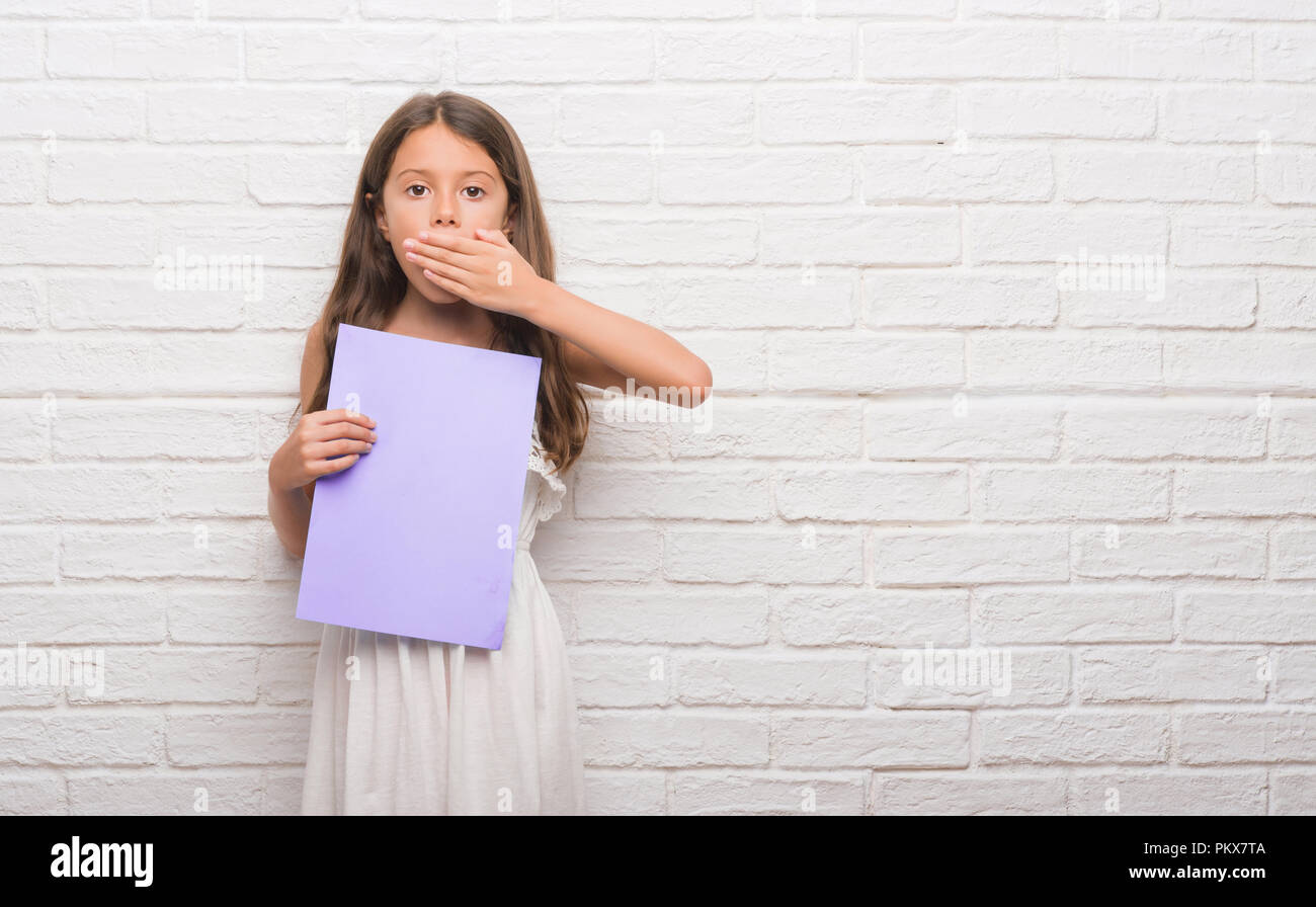 Young hispanic kid over white brick wall holding pink paper sheet cover ...