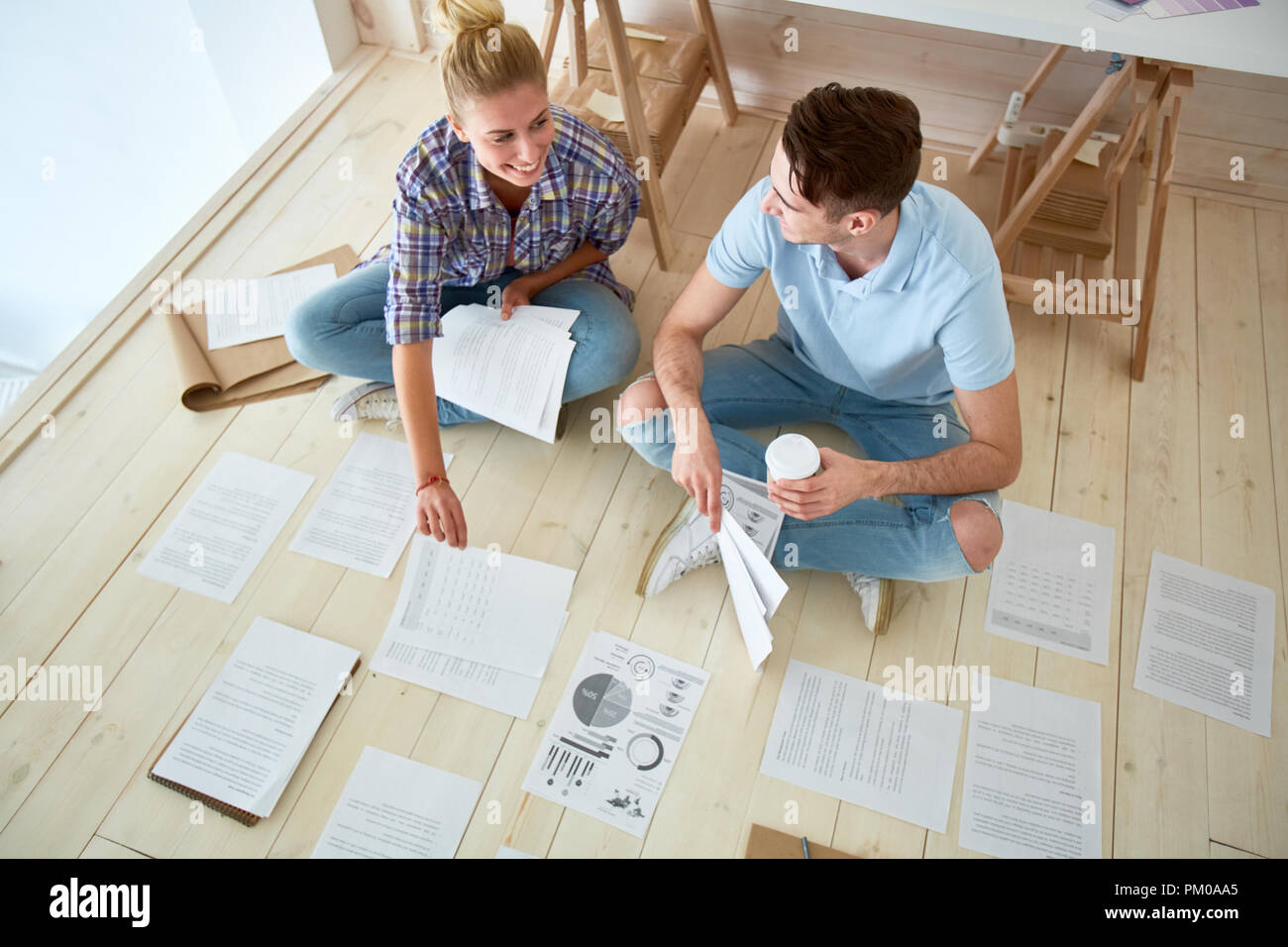 Two young employees in casualwear sitting on wooden floor of modern studio, working with papers ...