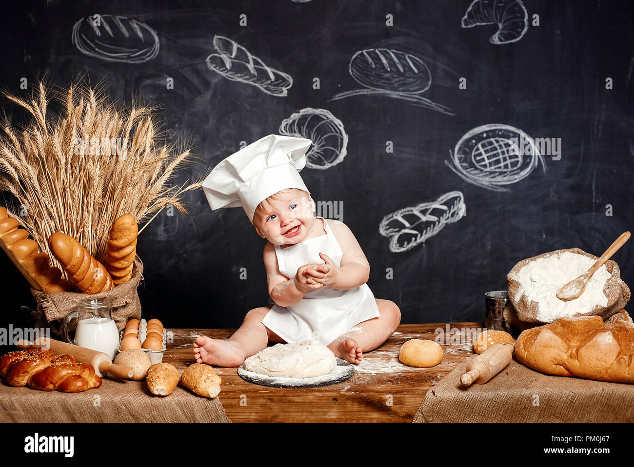 Charming toddler baby in hat of cook and apron sitting on table with ...