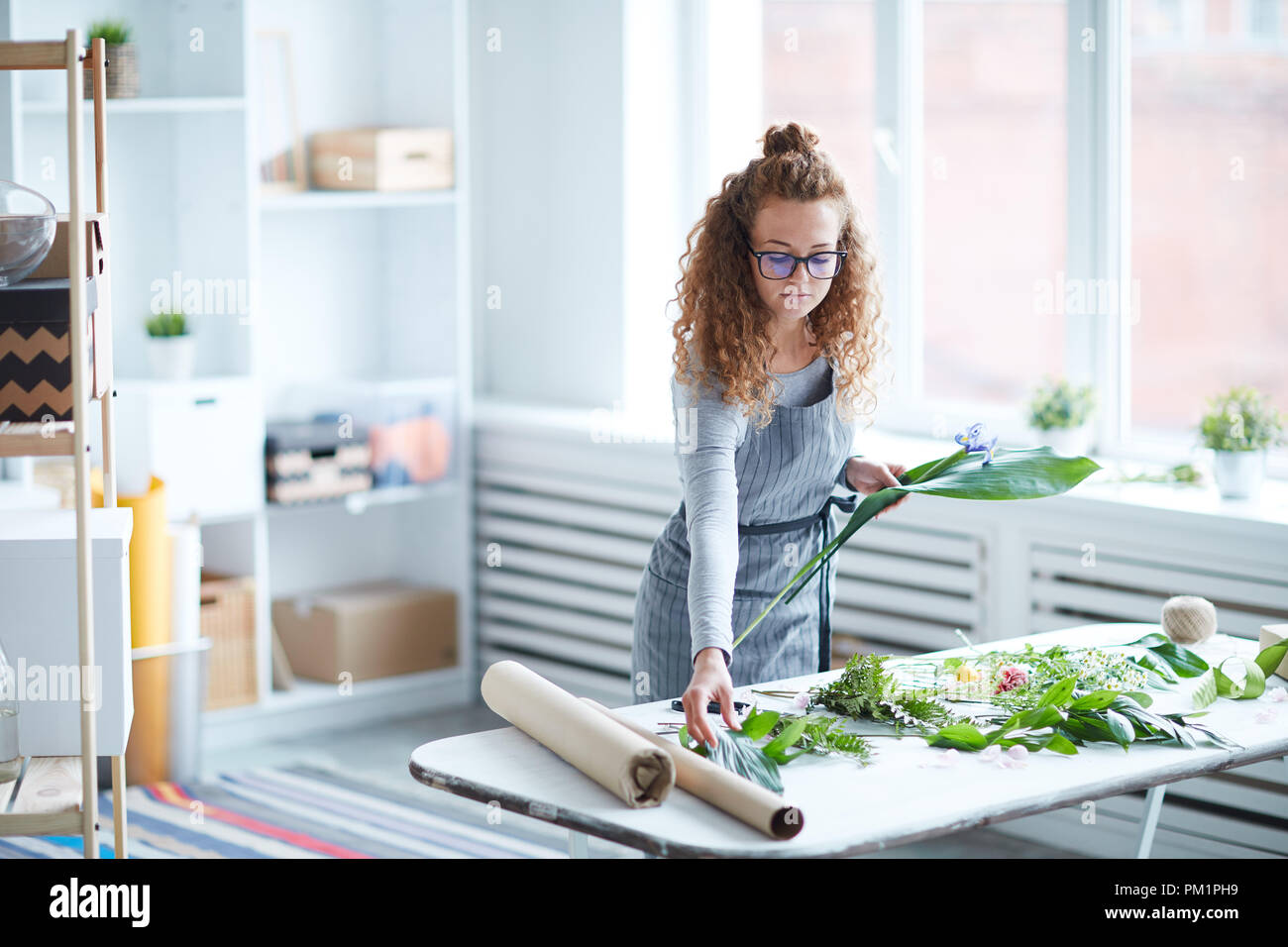 Young female florist leaning over workplace while arranging bunch of ...